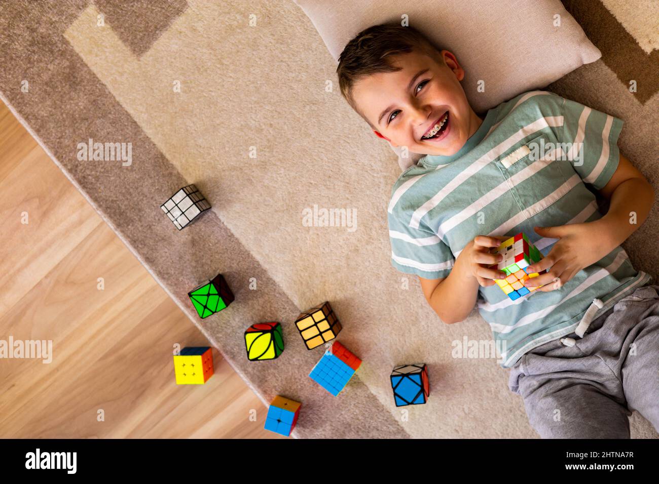 Relaxed adolescent boy lying on wooden floor assembling Rubik's cube ...