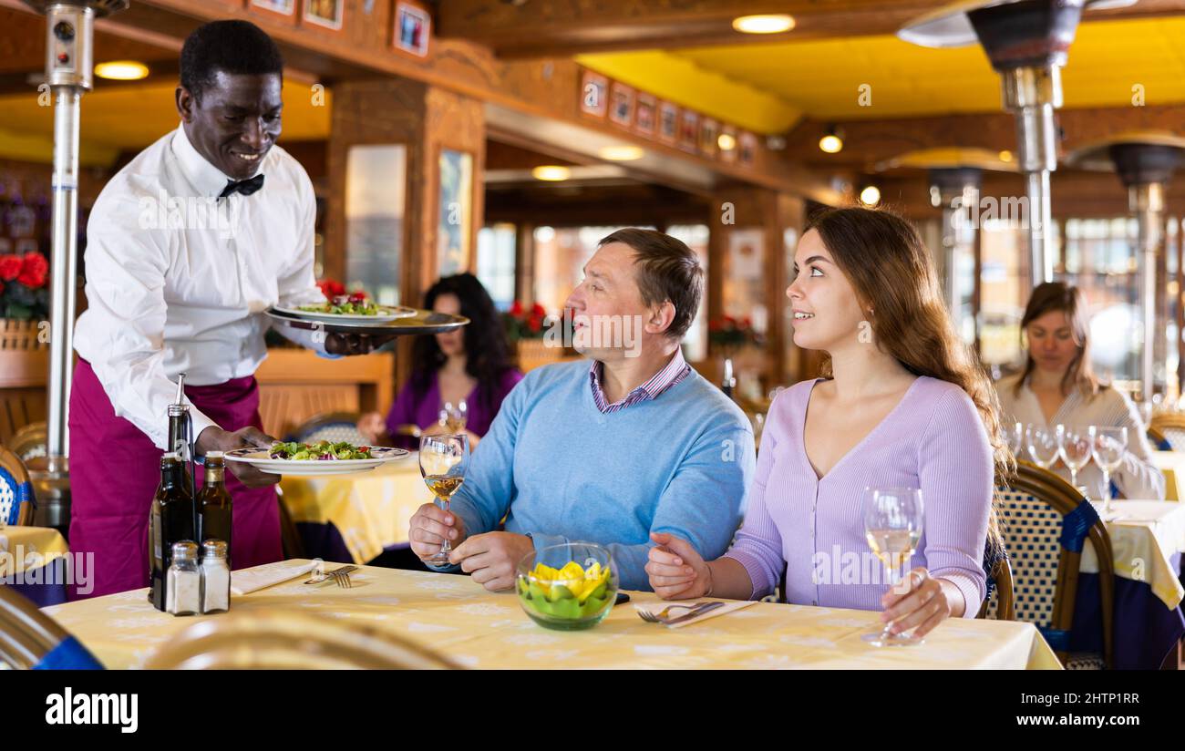 Polite african american waiter serving dishes to positive couple in ...