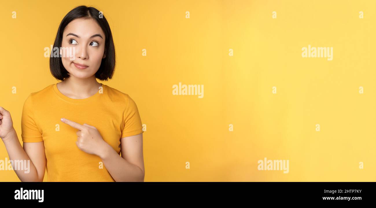 Portrait of intrigued asian woman, looking and pointing fingers left at ...