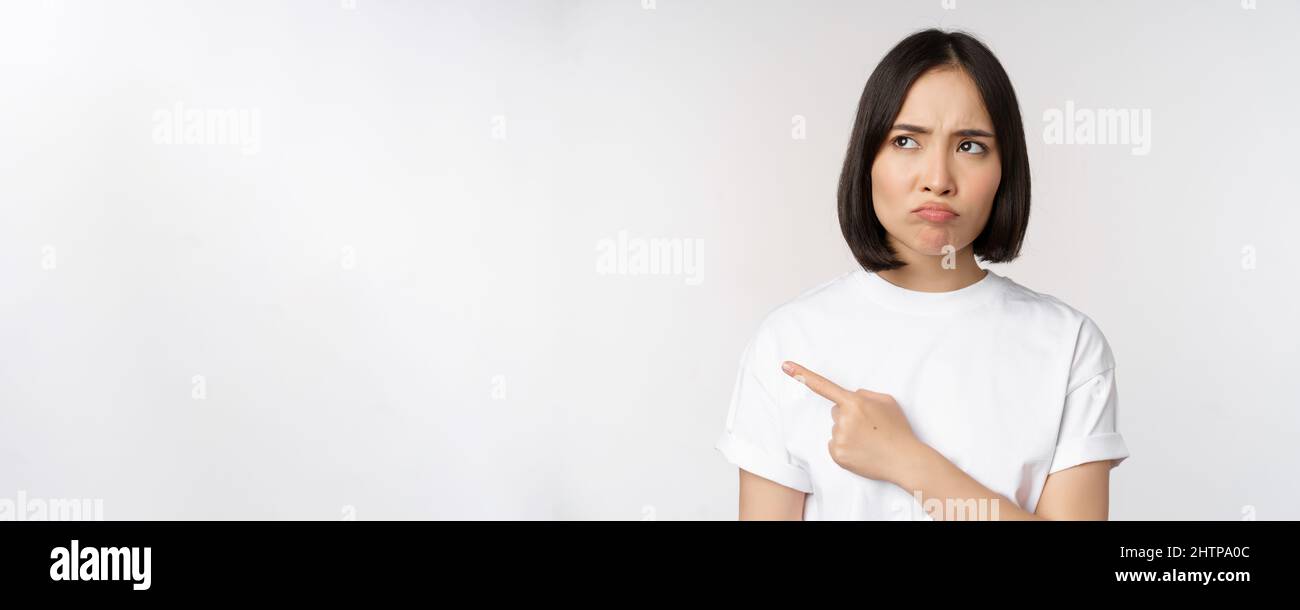 Angry and upset young asian woman pointing left, looking with regret or ...