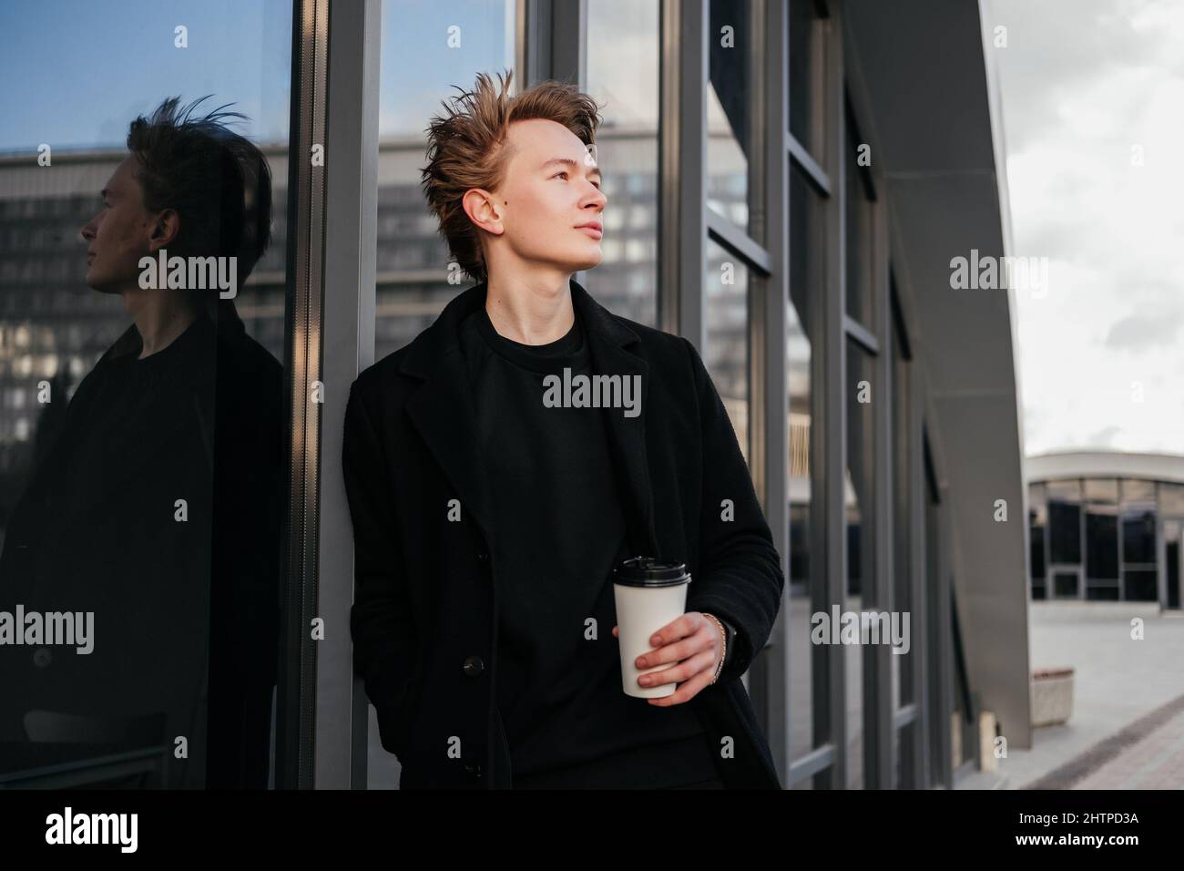 Thinking young man in black coat standing with cup of coffee in hands ...