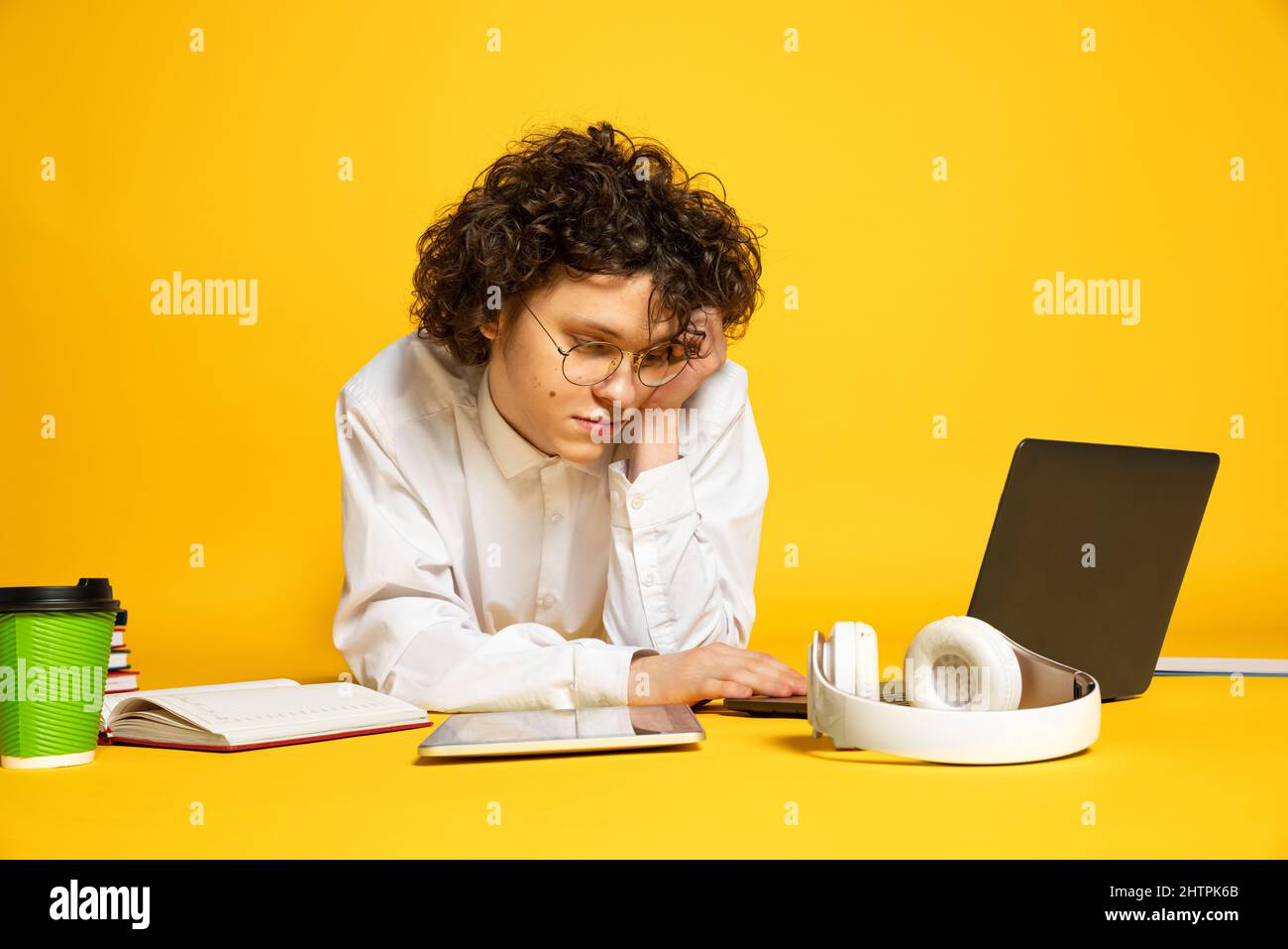 Portrait of young man, studnt studying, making homework on laptop isolated over yellow studio ...