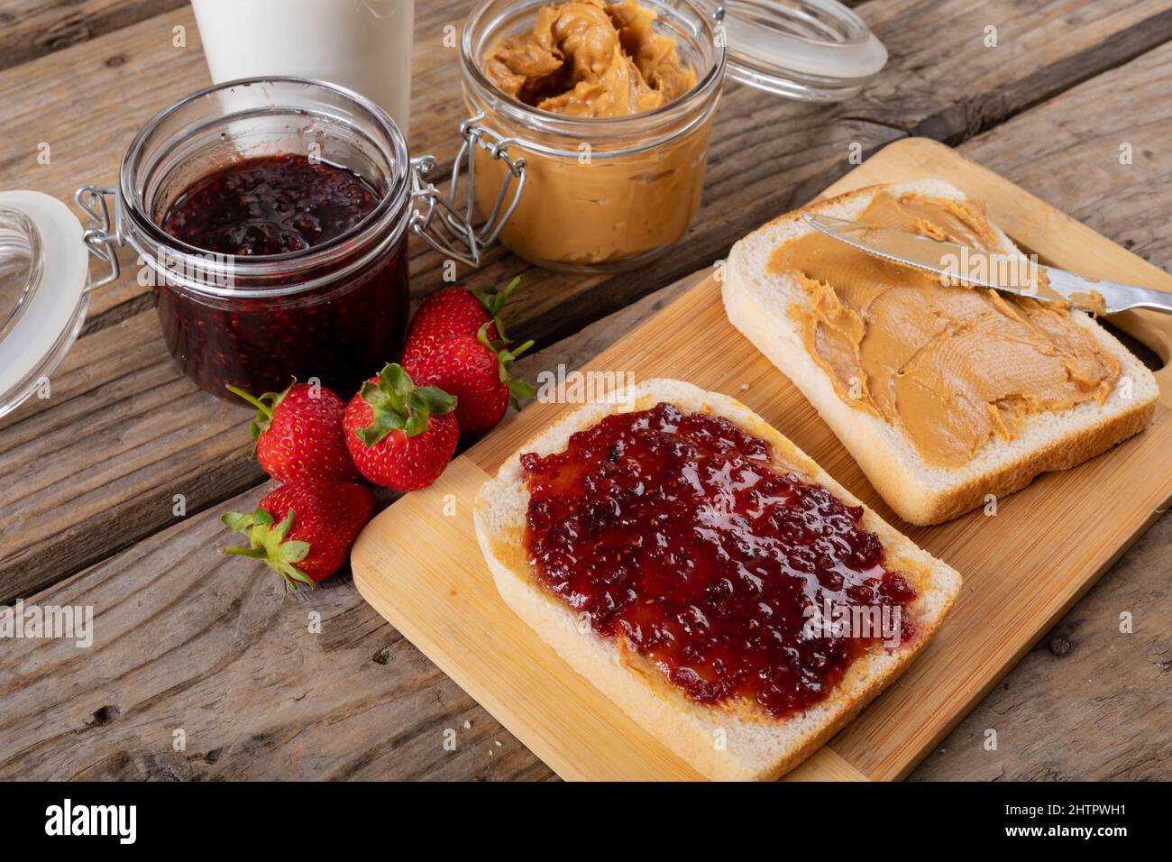 Close-up peanut butter and jelly sandwich on serving board with jars, strawberries and milk at ...