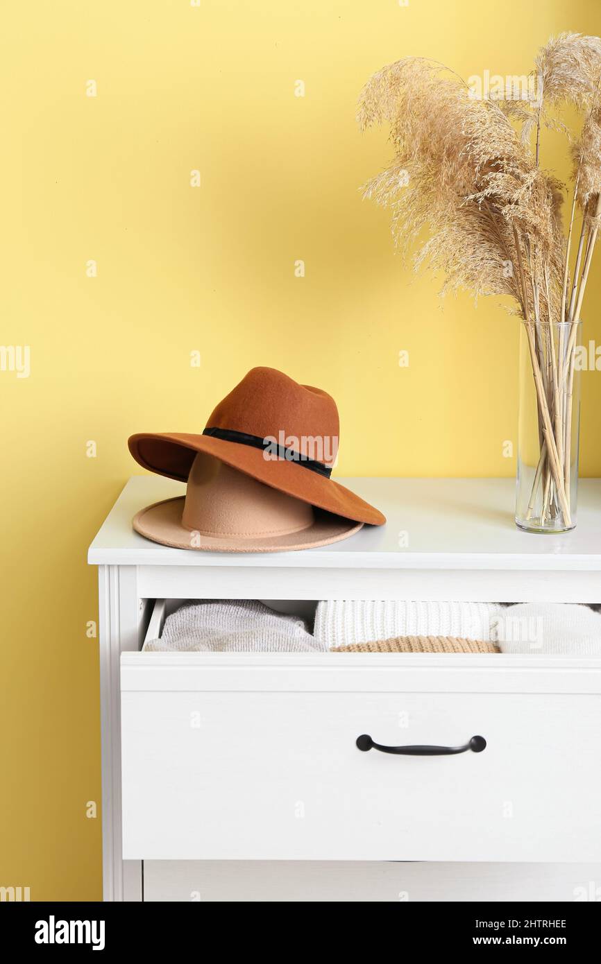 Chest of drawers with warm clothes, hats and vase near yellow wall ...