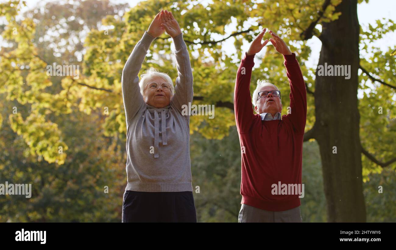 two caucasian elderly people exercising together in a park, trying to ...