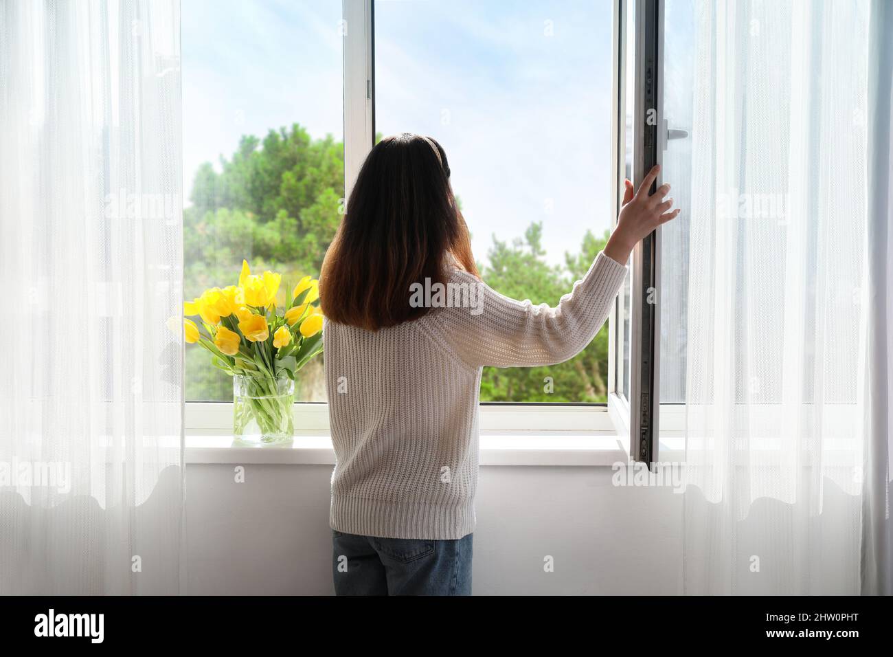 Pretty young Asian woman opening window at home on sunny day, back view ...
