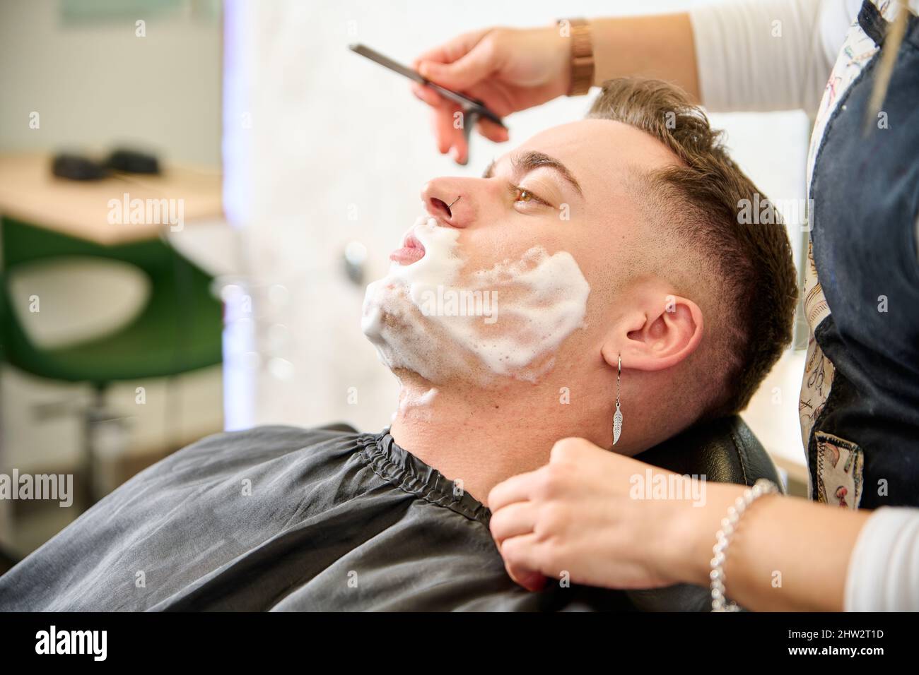Young man shaving with straight edge razor by hairdresser in barbershop ...