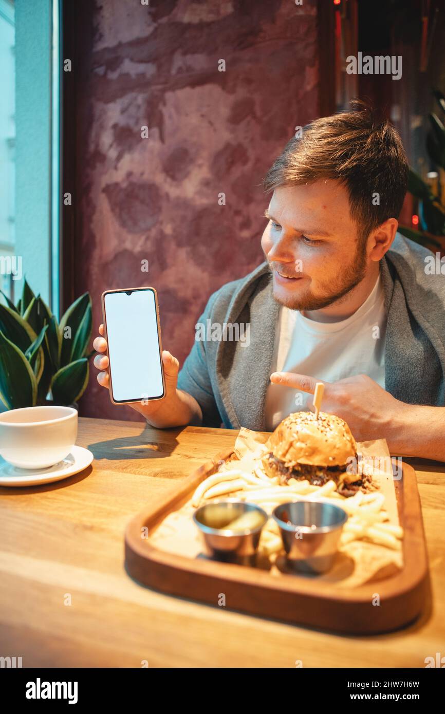 man in cafe holding phone with white screen eating burger drinking tea ...