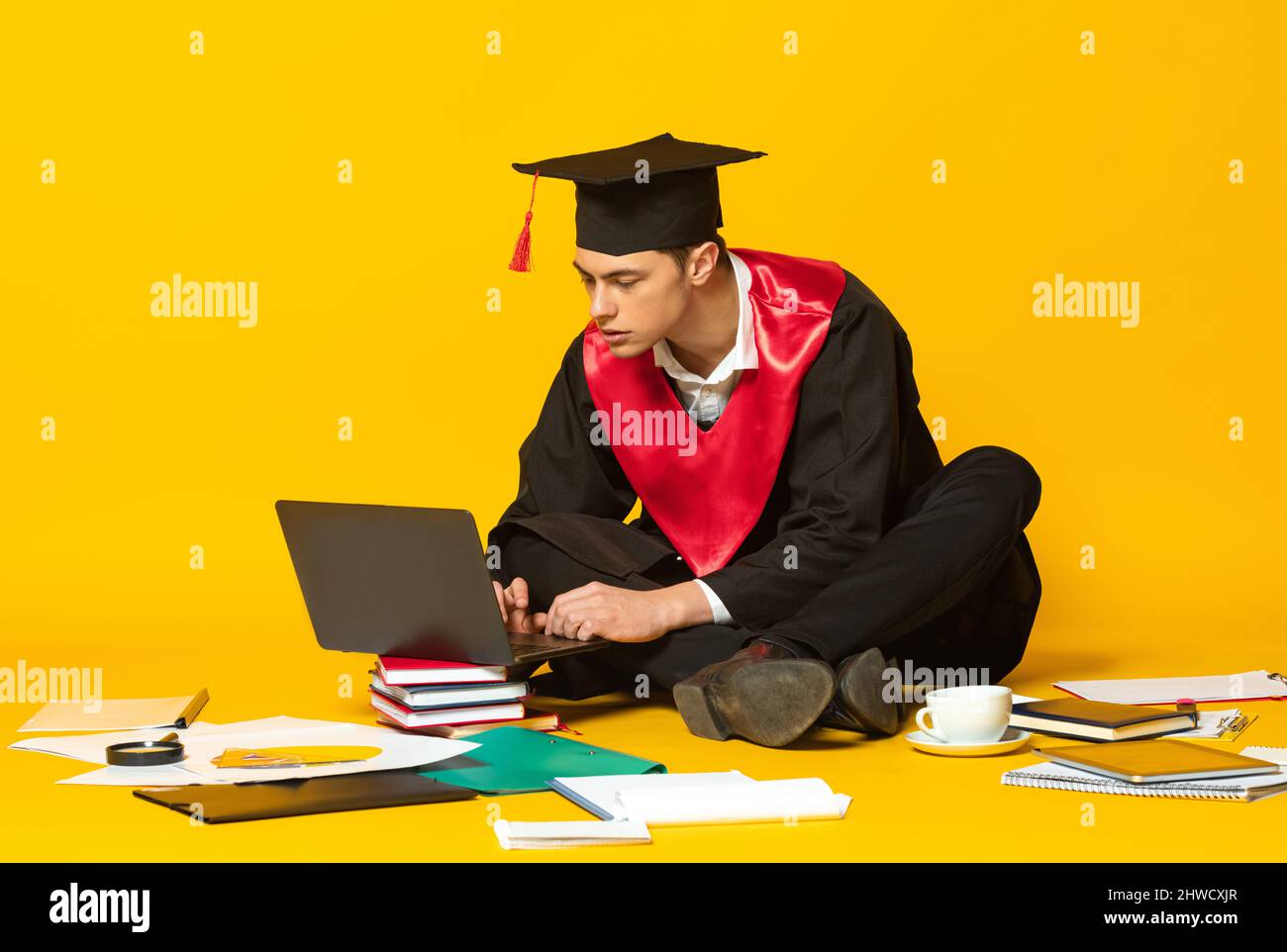 Portrait of young man, student in graduation cap and gown typing ...