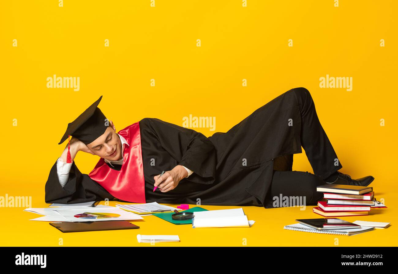Portrait of young man in graduation cap and gown lying on floor aroung ...