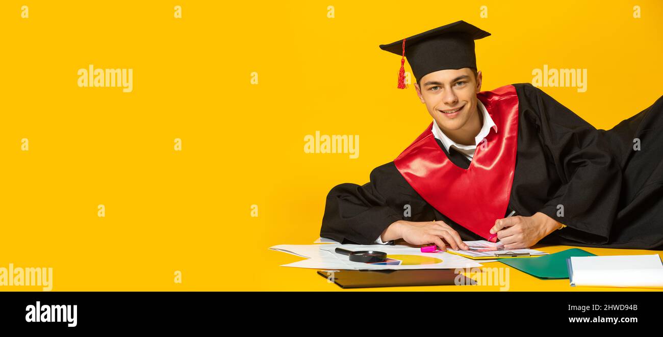 Portrait of young man in graduation cap and gown lying on floor aroung ...