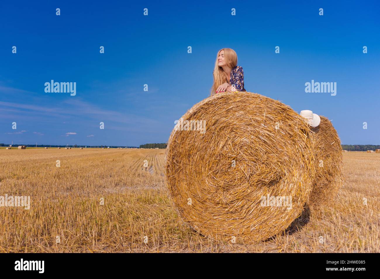 A blonde girl with long hair in a white hat is resting and posing near the sheaves of hay in a ...