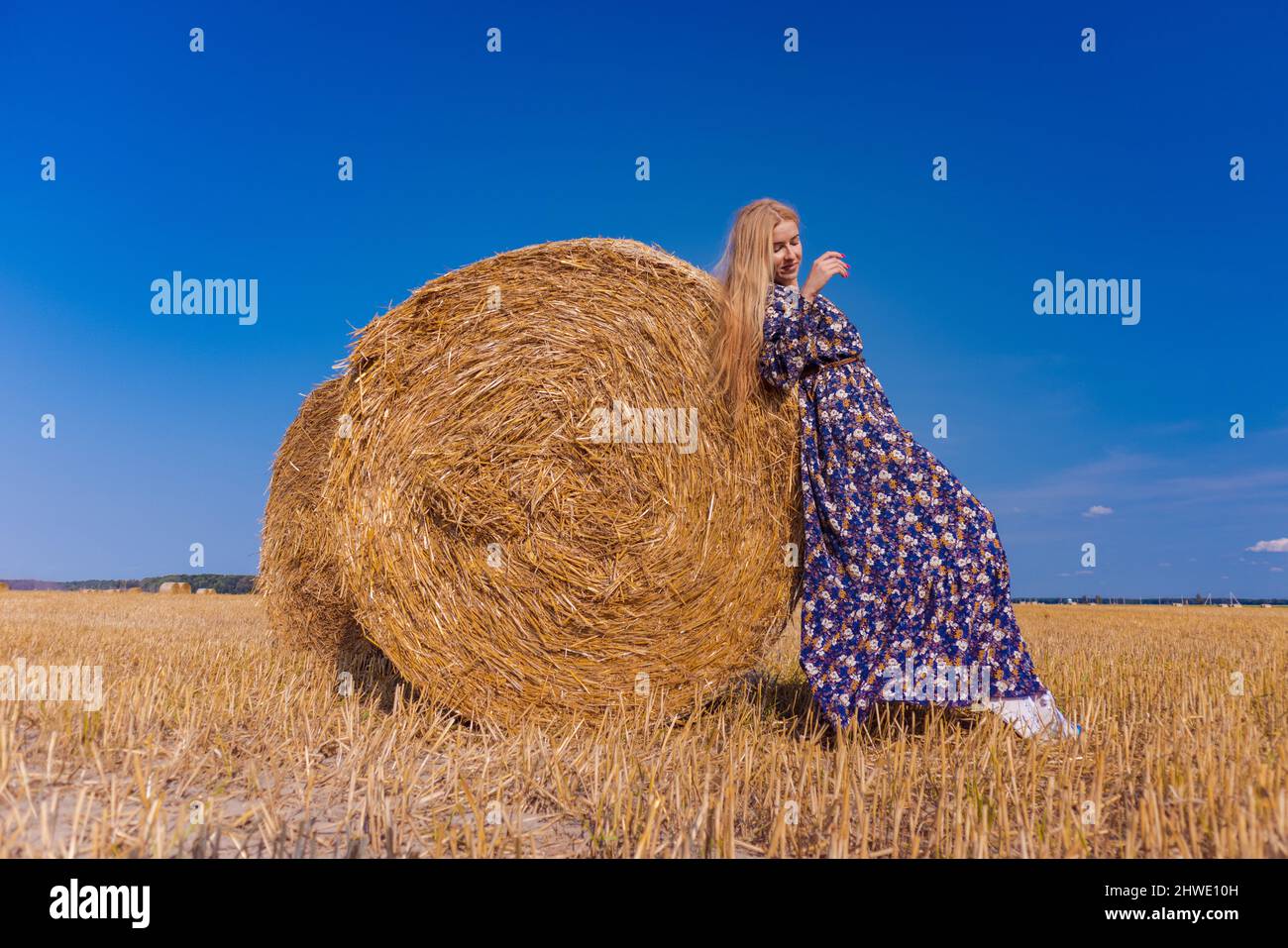 A blonde girl with long hair in a white hat is resting and posing near the sheaves of hay in a ...