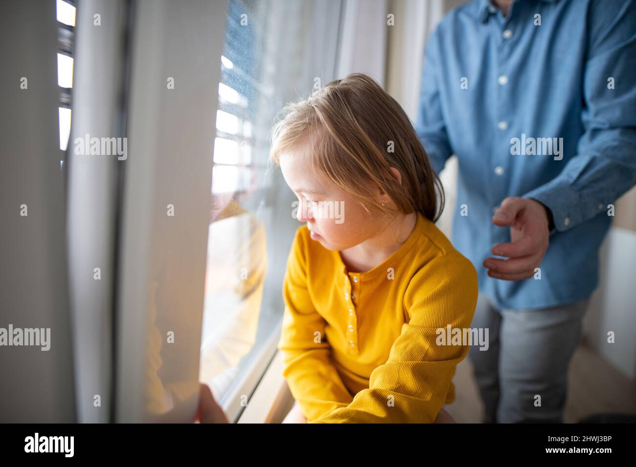 Sad little girl with Down syndrome looking throug window with her ...