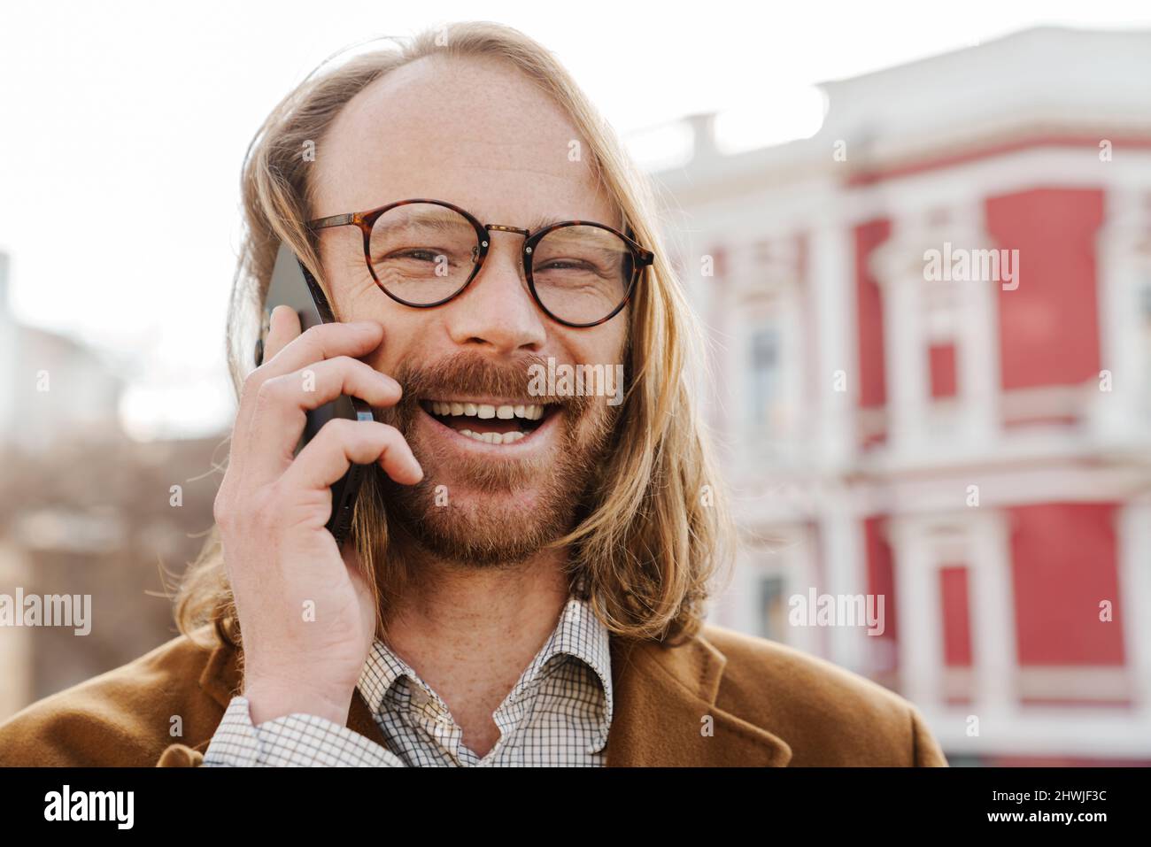 Ginger man in eyeglasses laughing while talking on mobile phone on city ...