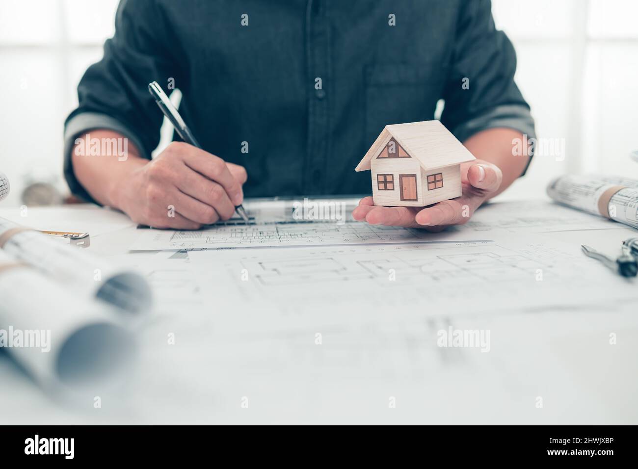 Architect engineer hands hold and show mini house model and use pen drawing on bueprint. House ...