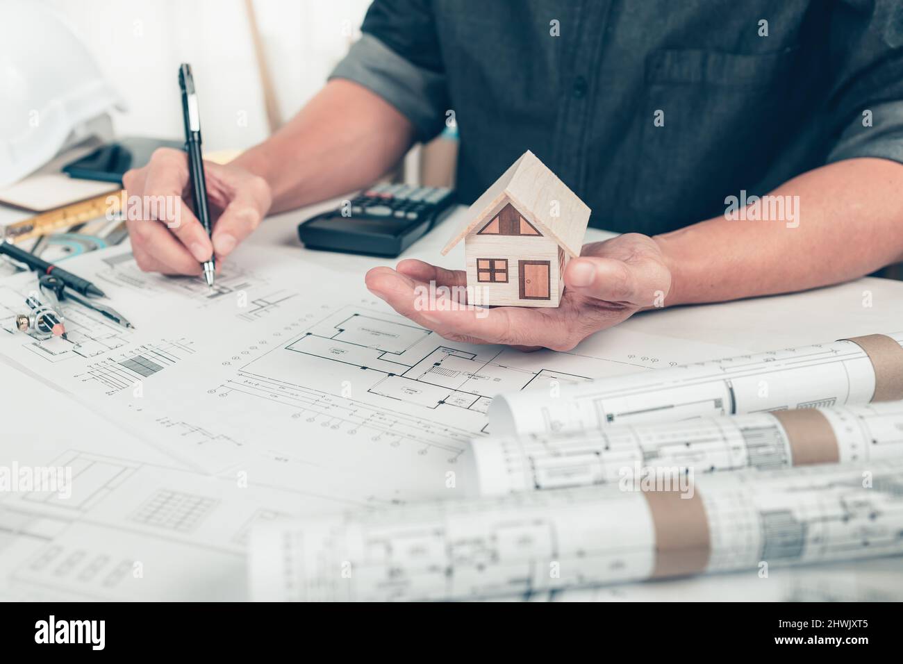 Architect engineer hands hold and show mini house model and use pen drawing on bueprint. House ...