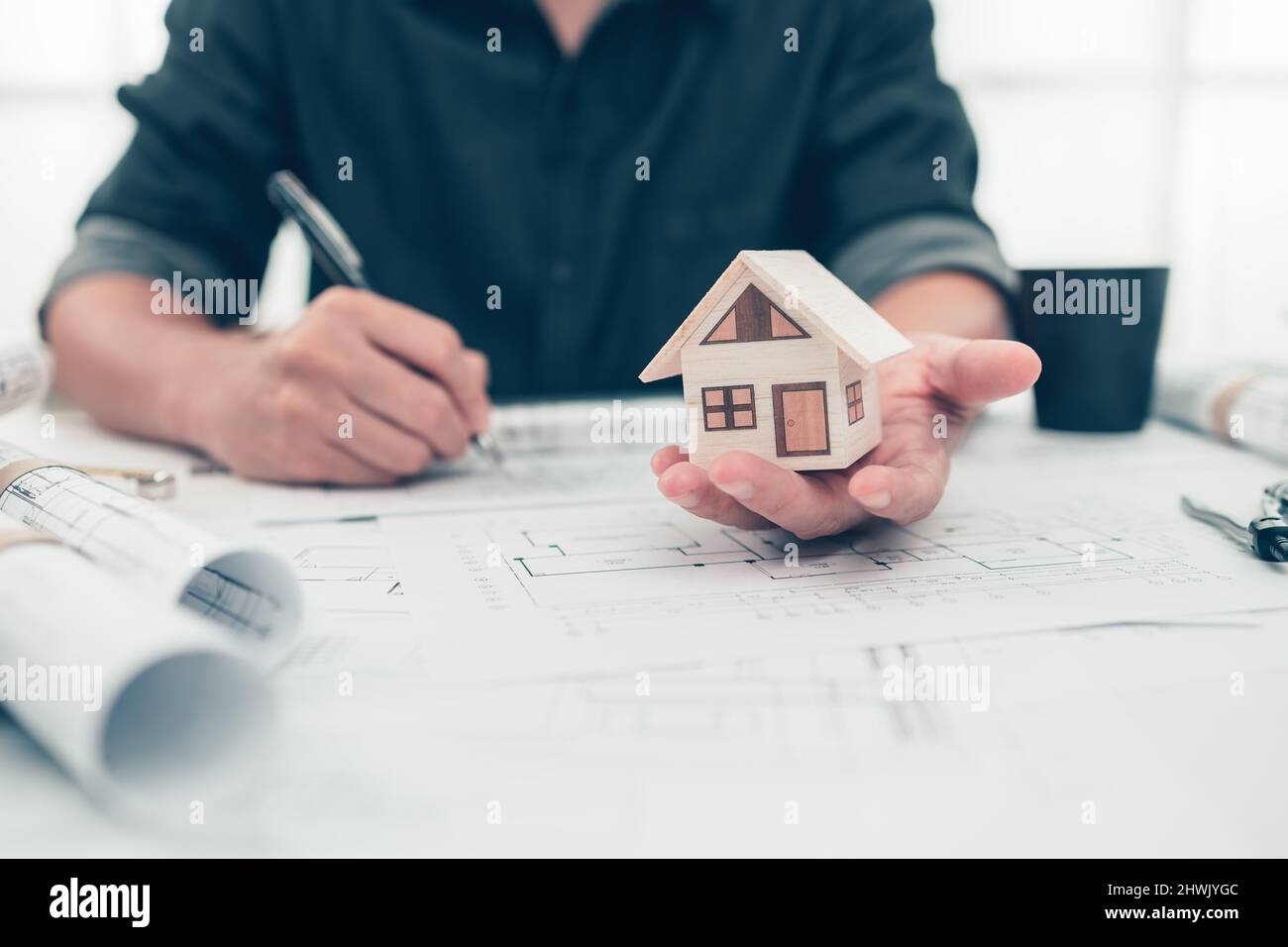 Architect engineer hands hold and show mini house model and use pen drawing on bueprint. House ...