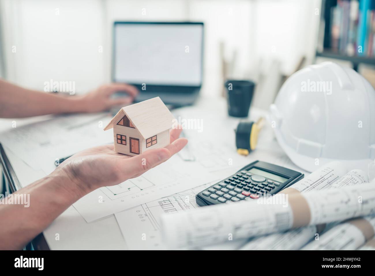 Architect engineer hands hold and show mini house model and use laptop on desk. House planning ...