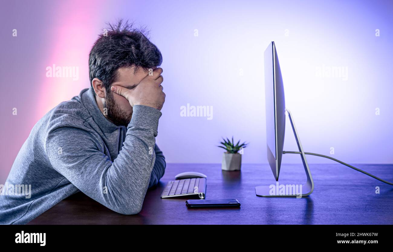 A tired man sits in front of a computer, covering his face with his ...