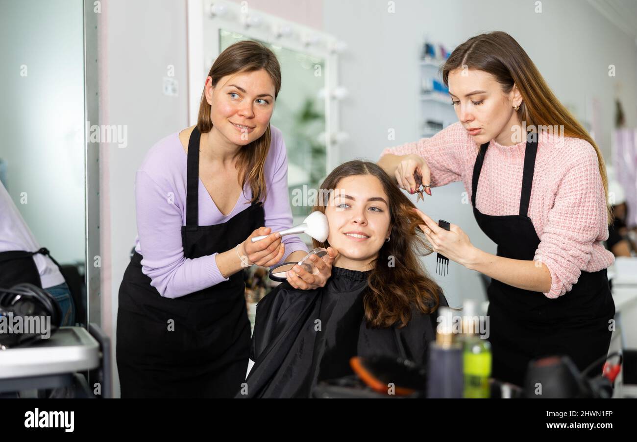 Professional female stylists making hairstyle and makeup to young woman ...