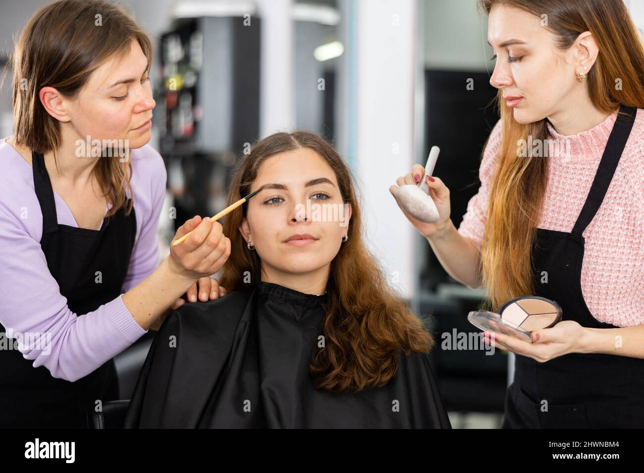 Two makeup artists applying decorative cosmetics to young female client ...