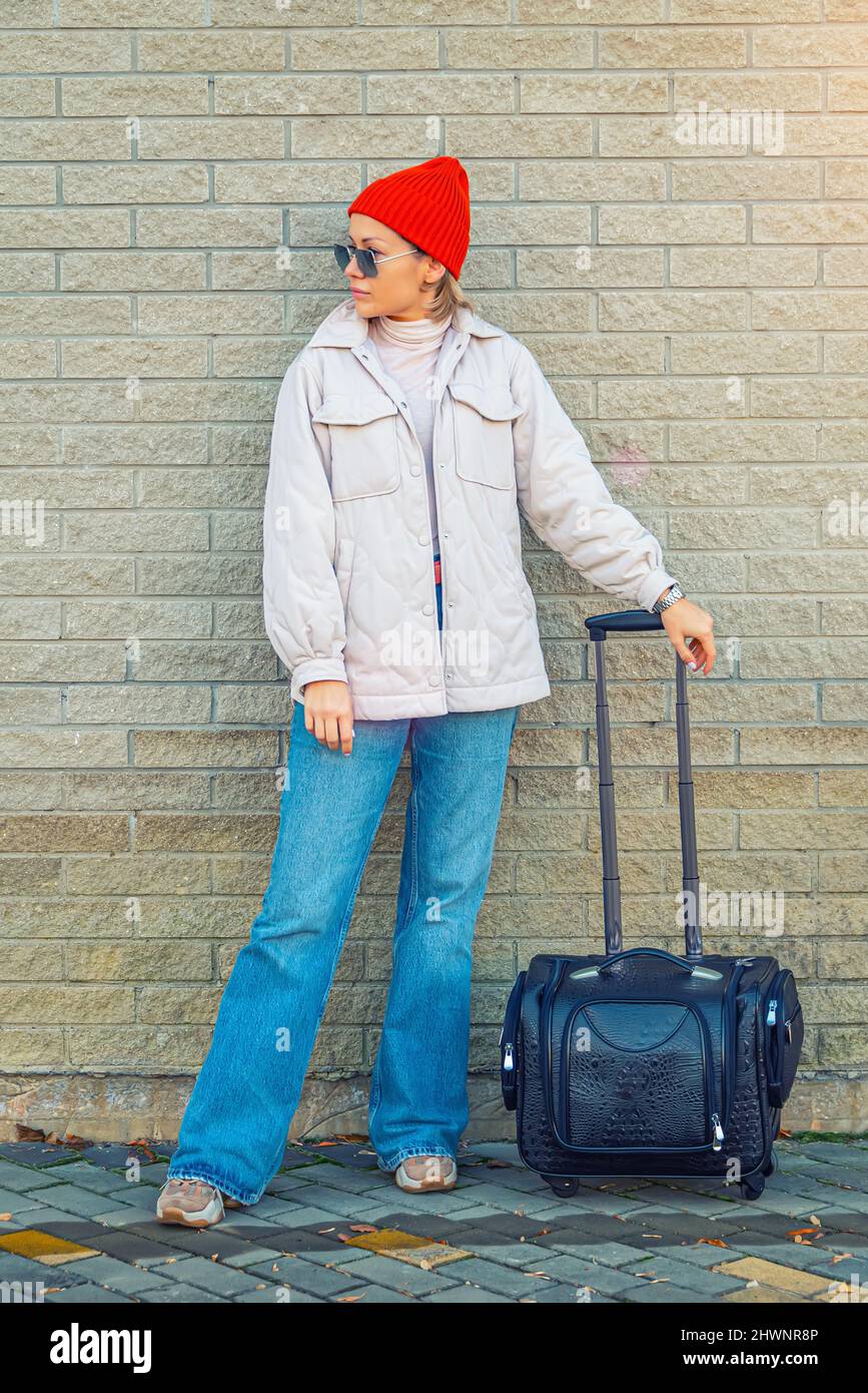 A young woman in casual clothes and with a suitcase poses at the ...