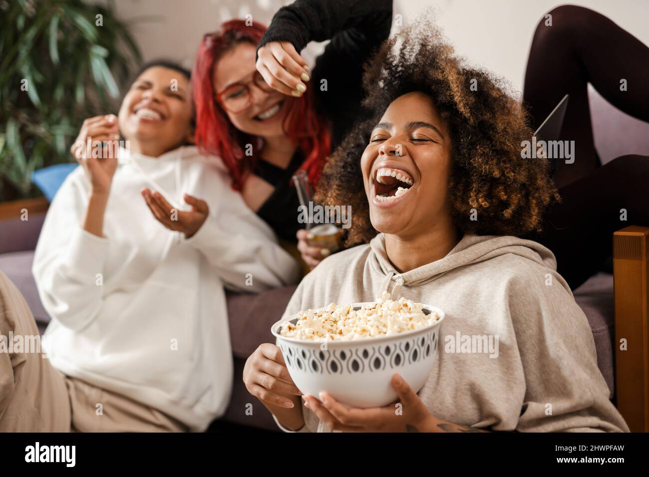 Three happy young mix race women friends sitting on a couch drinking ...