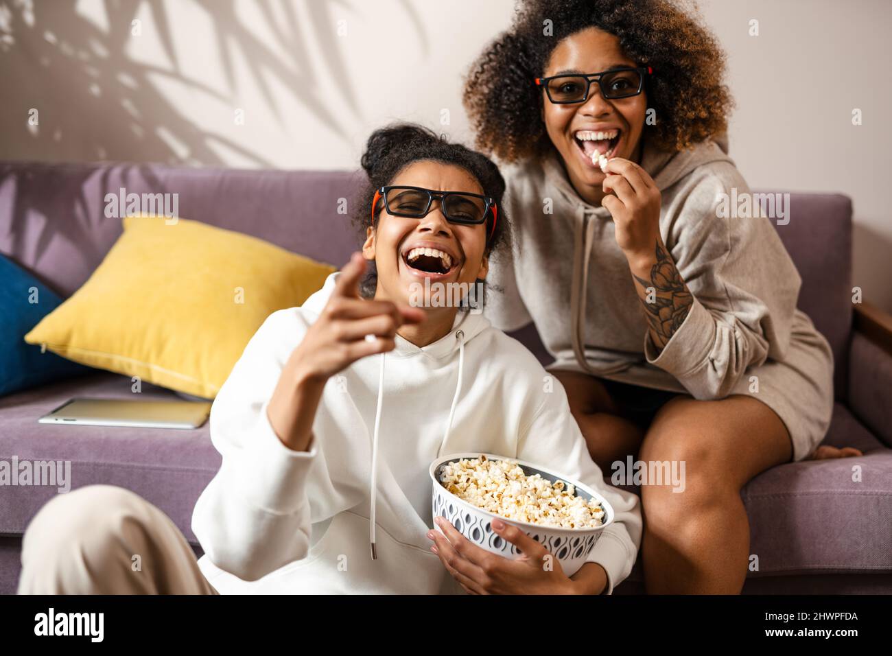 Two happy multiethnic women friends sitting on a couch at home with ...