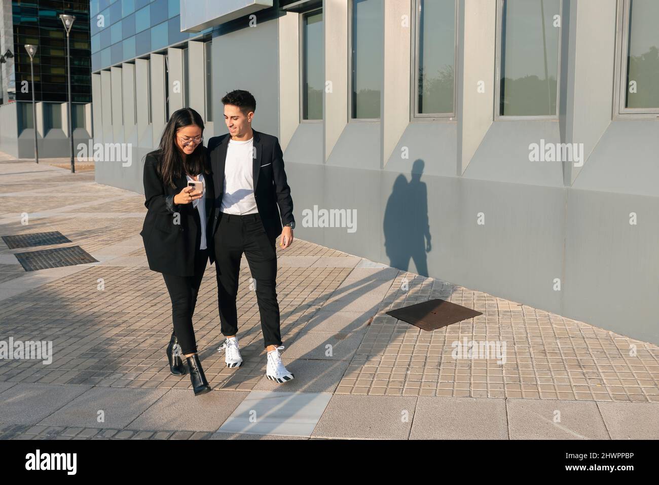 Business colleagues sharing smart phone walking on footpath by office ...
