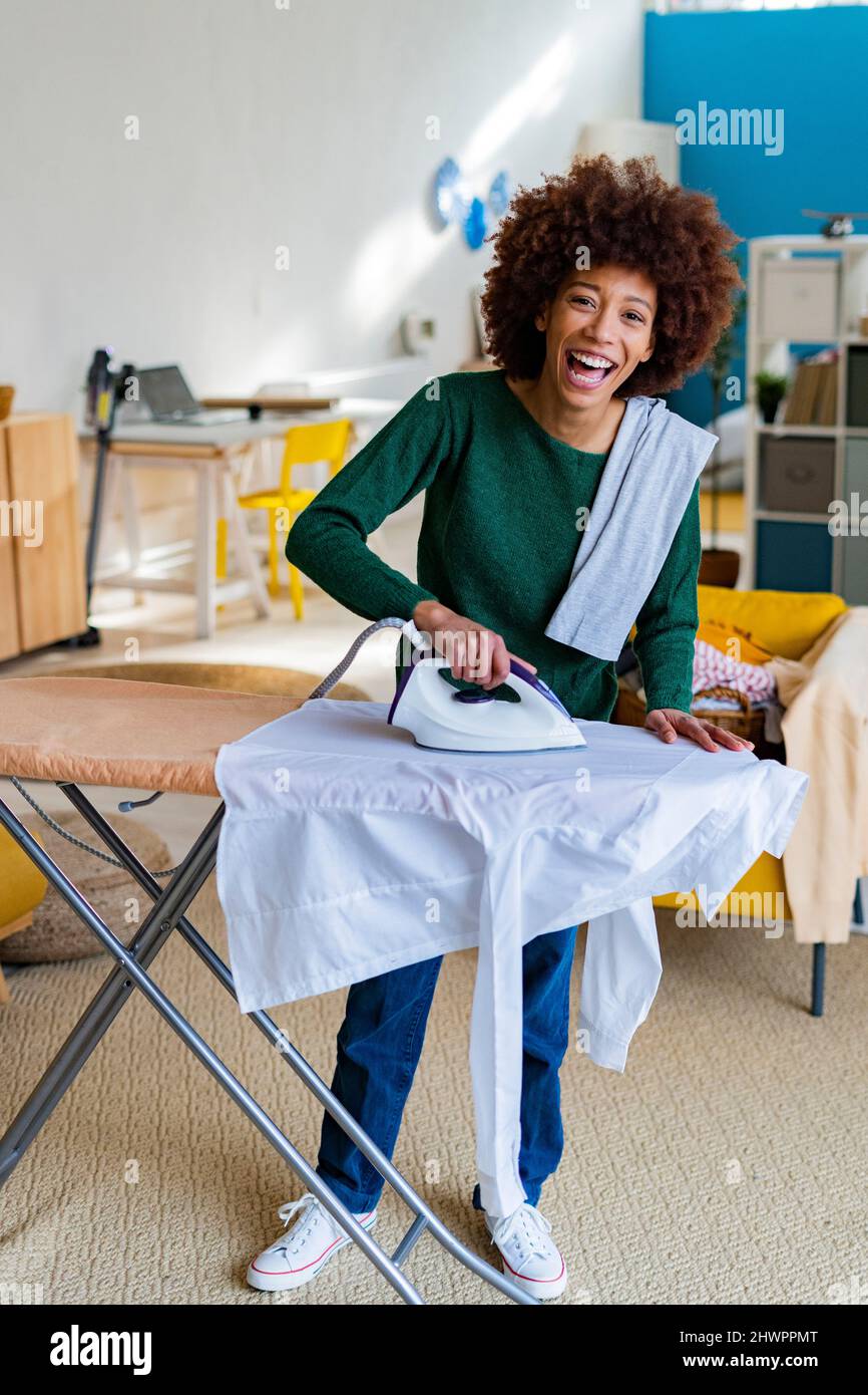 Cheerful young Afro woman ironing shirt on board in living room Stock ...