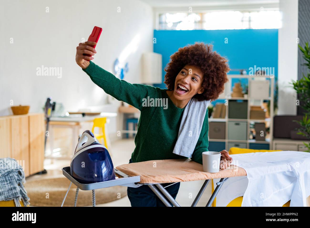 Cheerful young woman taking selfie with coffee mug at ironing board in ...