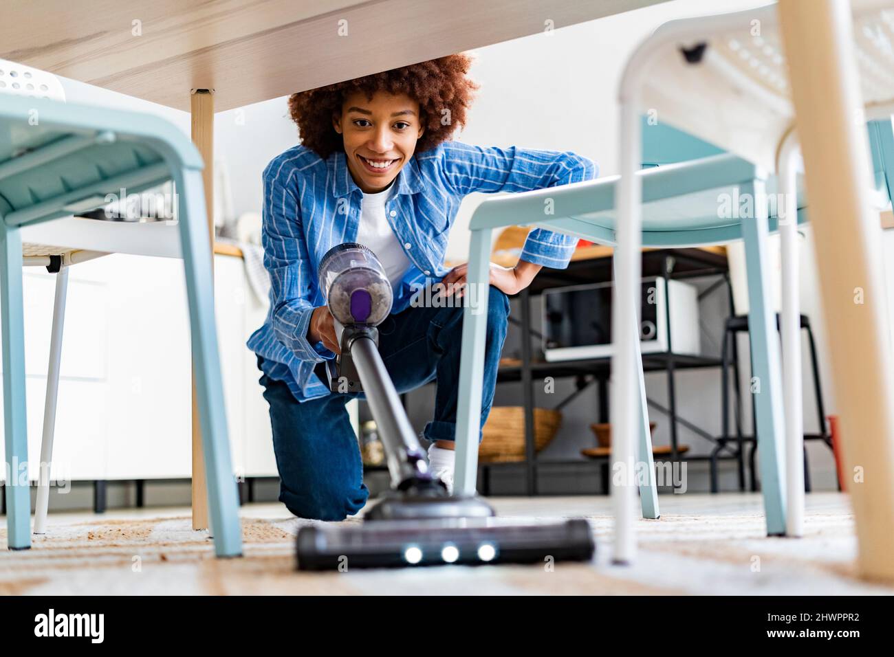 Smiling young woman crouching using vacuum cleaner under dining table ...