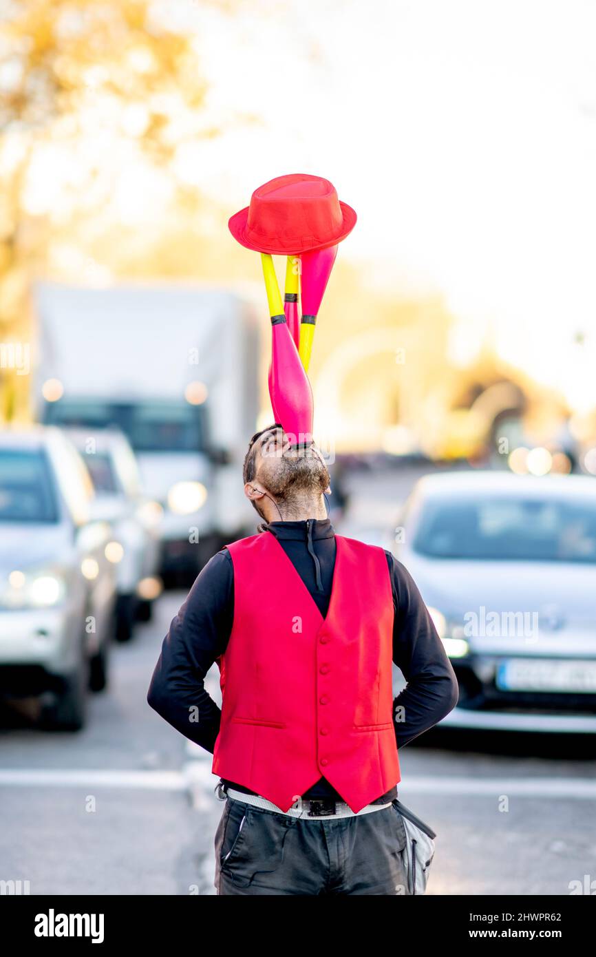 Performer balancing juggling pins and hat on nose in front of cars ...