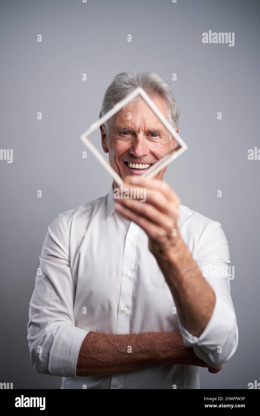 Smiling businessman looking through frame in front of gray background ...