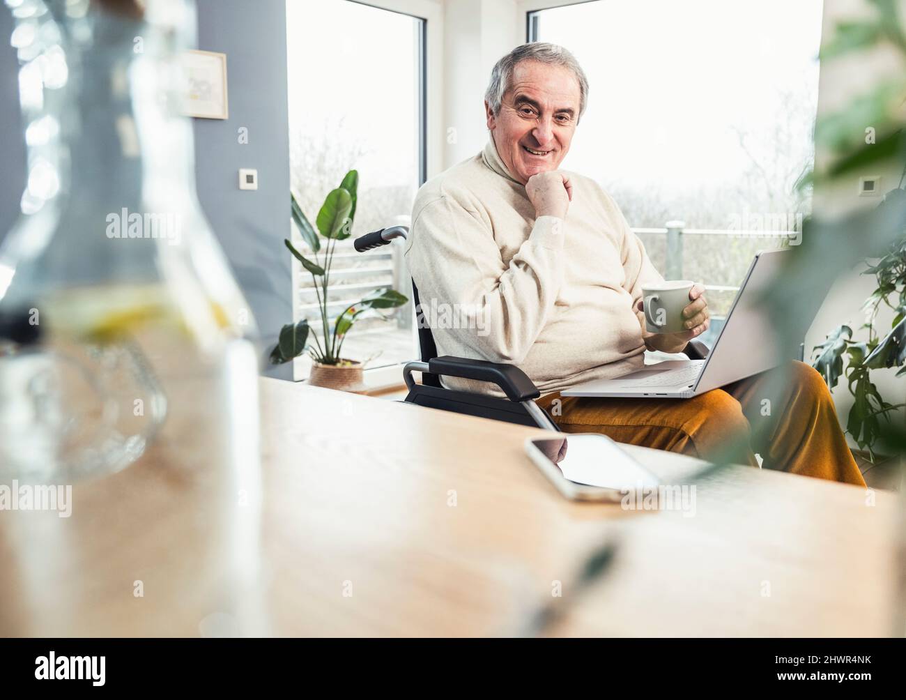 Smiling disabled man holding coffee cup sitting with hand on chin at ...