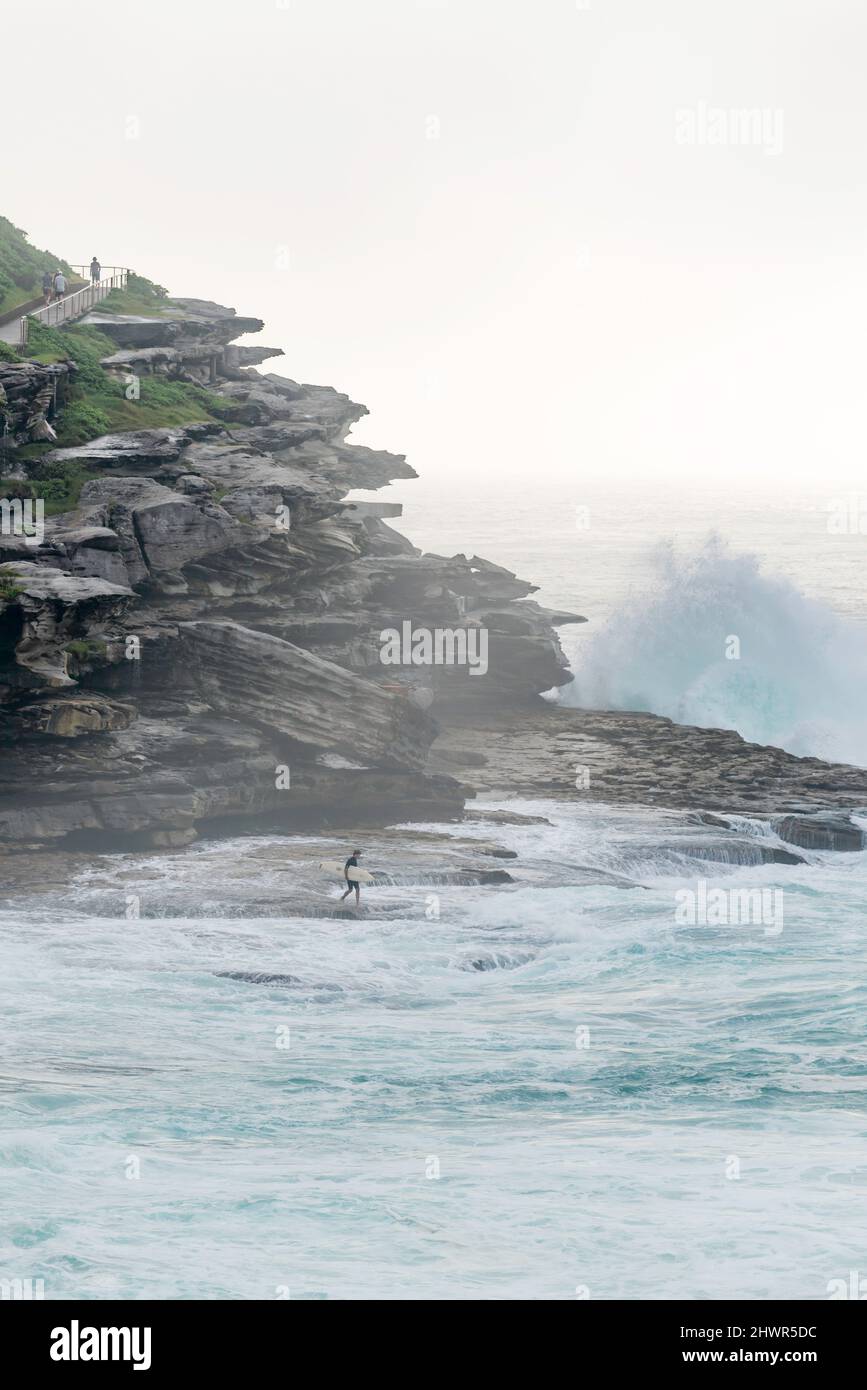 A lone board rider enters the water as big waves crash on the rocky ...