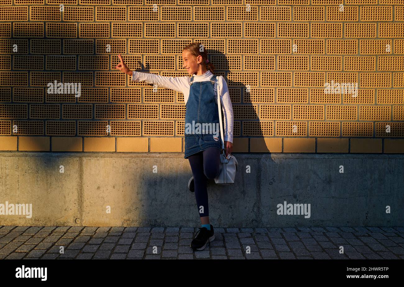 Happy girl gesturing peace sign standing on one leg in front of brick ...