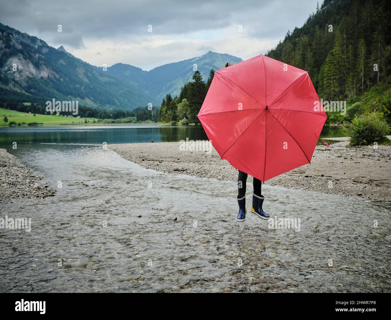 Girl covering face behind red umbrella standing on water at lakeshore ...