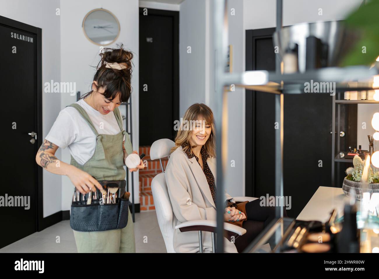 Happy make-up artist talking with customer sitting on chair at salon ...