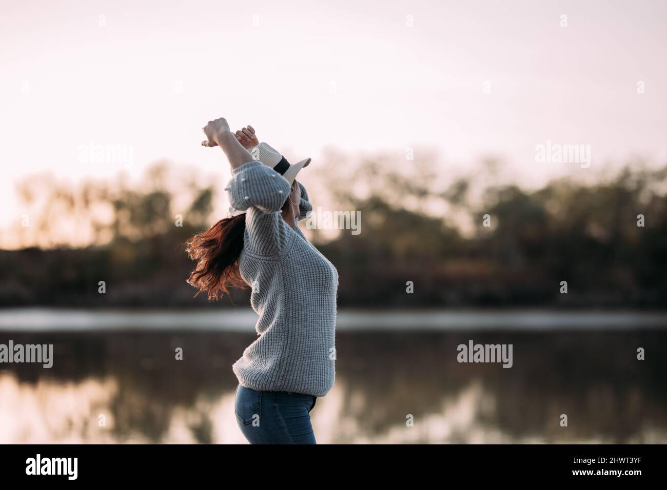 Brunette woman dancing happily on the shore of a river with a hat ...
