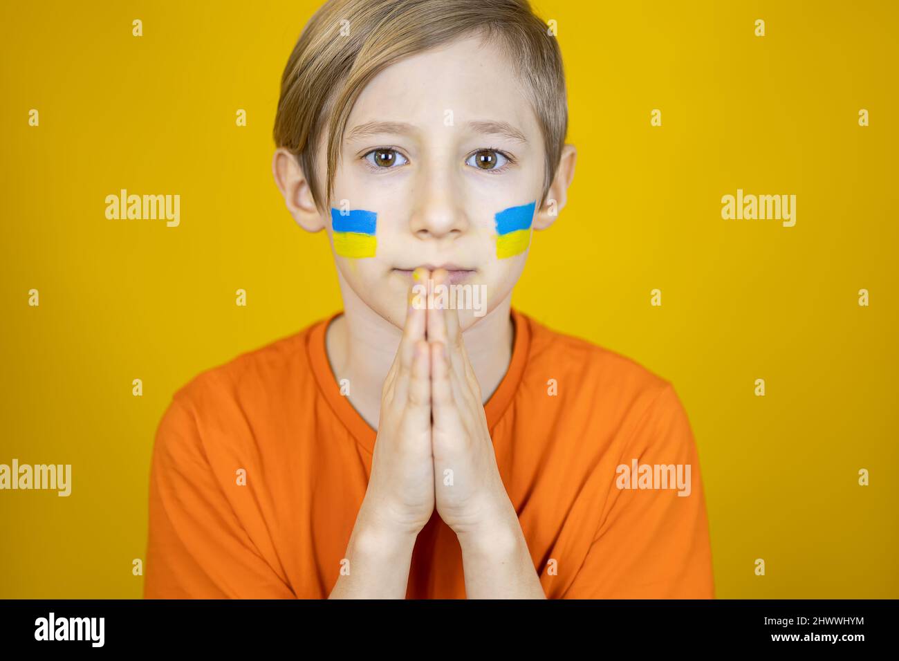 a sad boy with a painted flag of Ukraine on his cheeks folded his palms ...