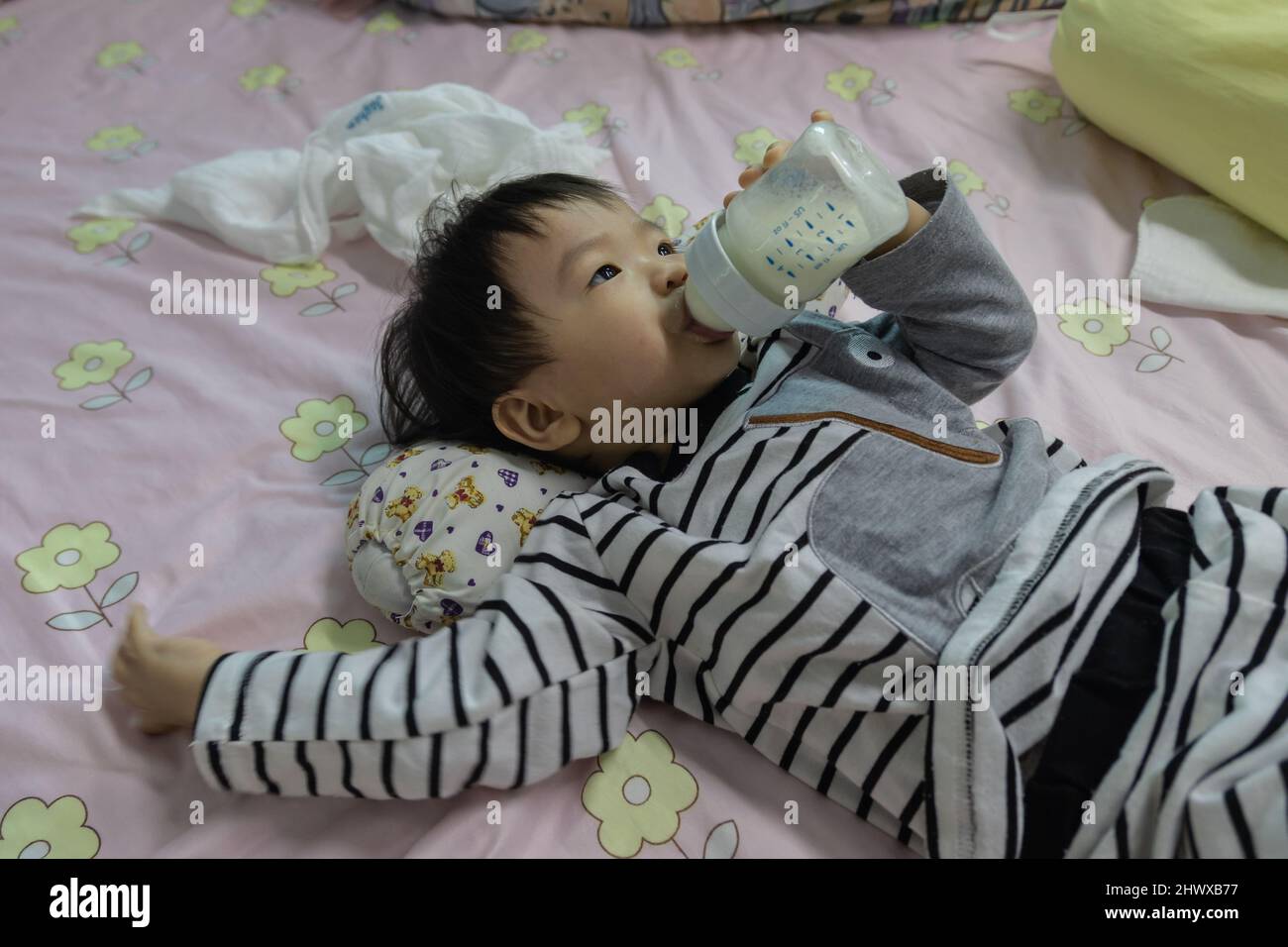 Asian Chinese baby boy lying on bed with holding a milk bottle drinking ...