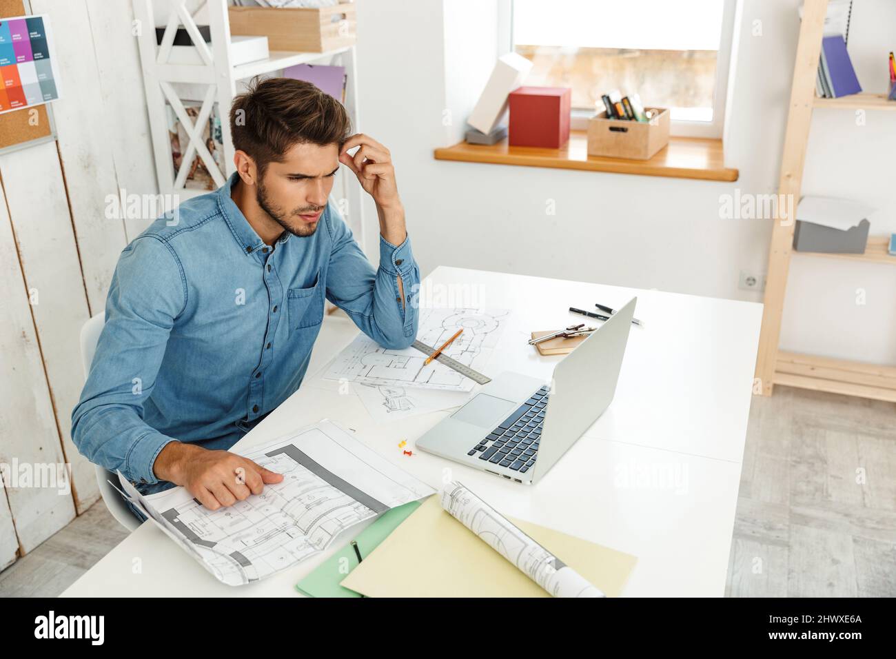 Young white man working with laptop and drawings in office indoors ...