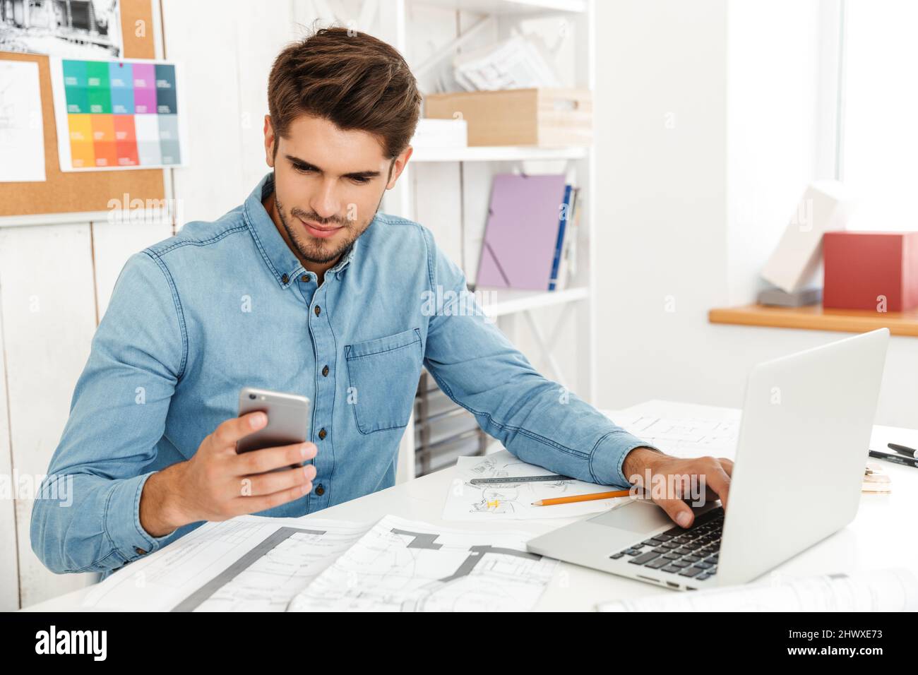 Young man using cellphone while working with laptop and drawings in ...