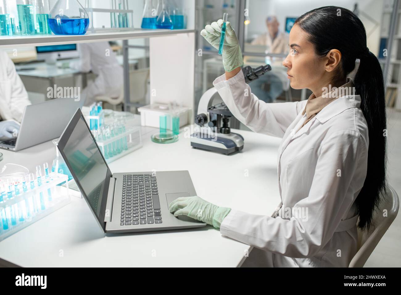 Young serious female lab worker in whitecoat sitting by workplace in ...