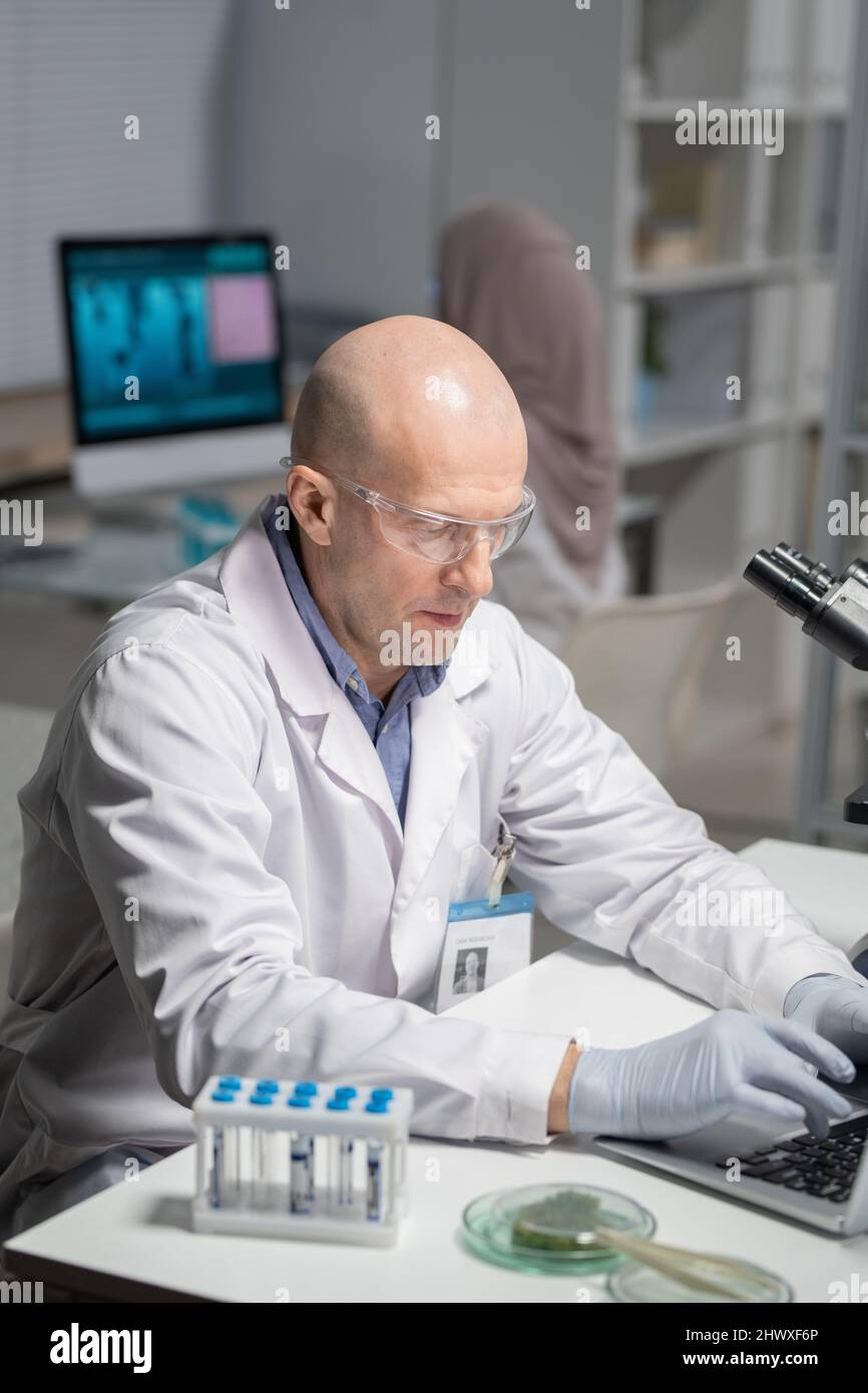 Mature male in lab coat and protective gloves and eyewear typing on ...