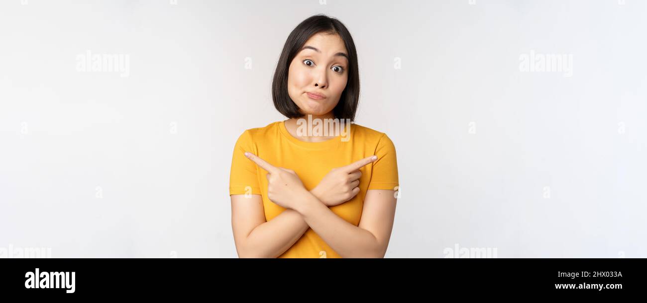 Complicated asian girl pointing fingers sideways, showing left and right choices, staring ...