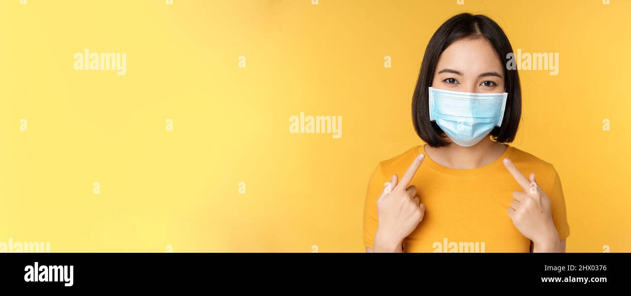 Portrait of smiling asian woman in medical face mask, pointing at her ...