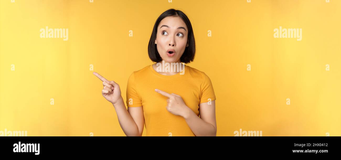 Portrait of intrigued asian woman, looking and pointing fingers left at ...